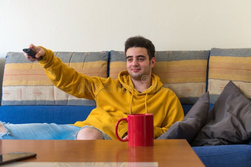 Young Man Relaxing Turning On The Tv Smiling Laying On The Sofa To
