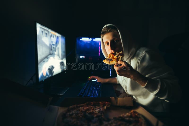 Young Man Sits at Night at a Table Next To a Computer, Eating a Slice ...
