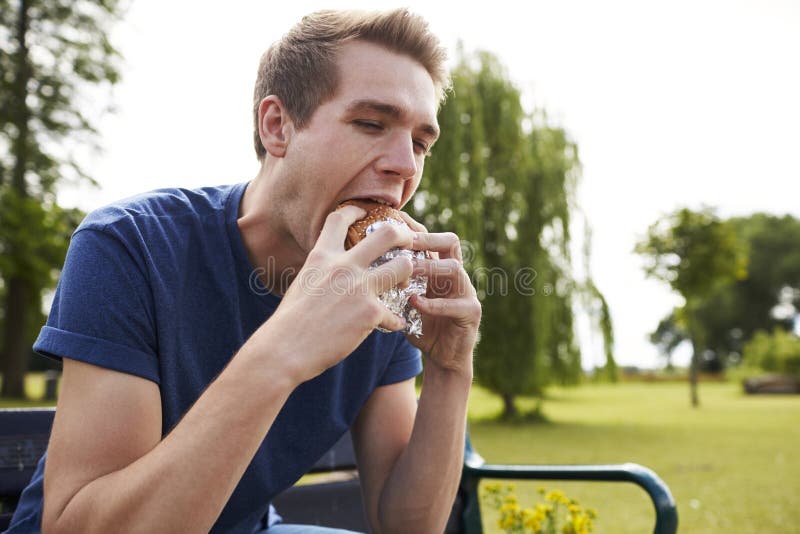 Young Man Sitting on Park Bench Eating Burger Stock Image - Image of ...