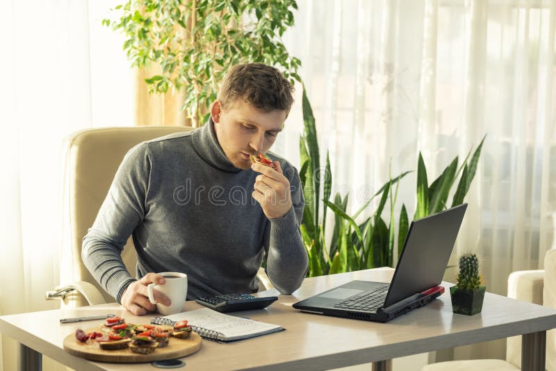 A Young Man Snacks on Sandwiches while Working at the Workplace. the ...
