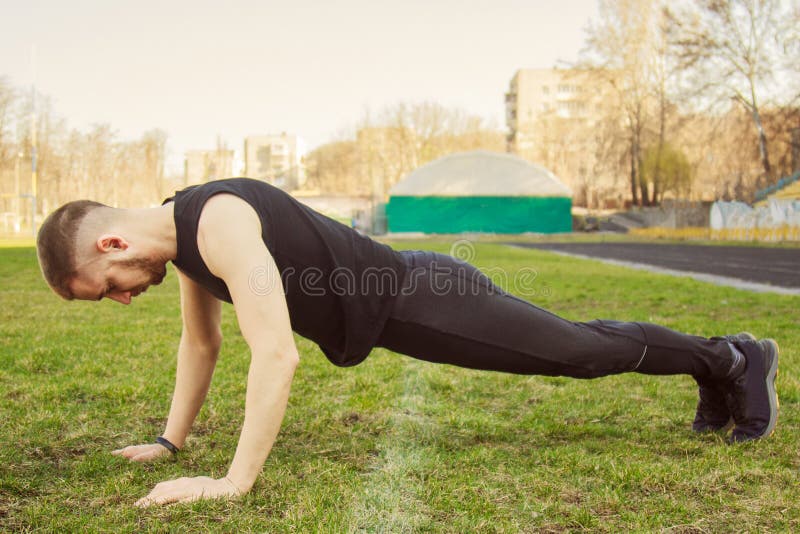 Young Man in Sportswear Does Exercises on the Grass at the Stadium ...