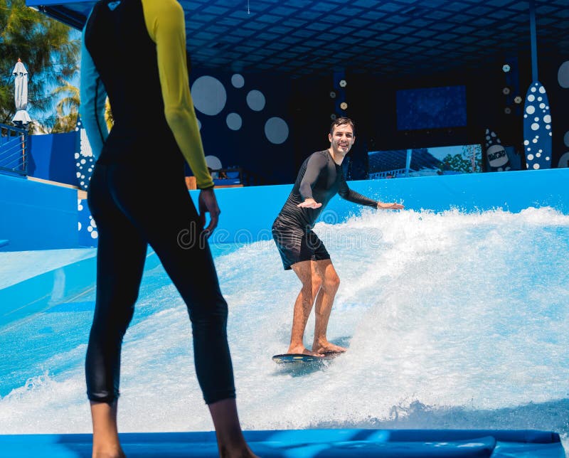 Young Man Surfing with Trainer on a Wave Simulator at a Water Amusement ...