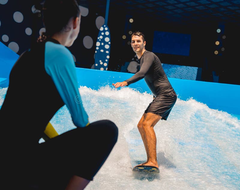 Young Man Surfing with Trainer on a Wave Simulator at a Water Amusement ...