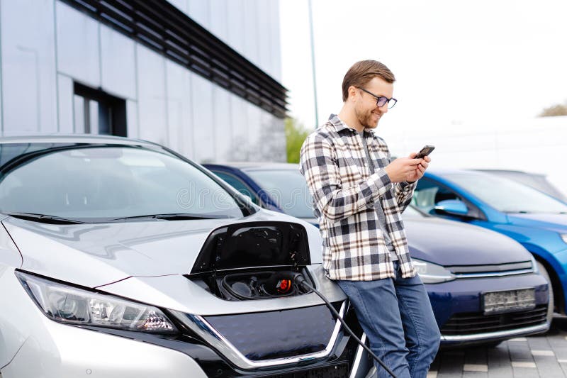 A Young Man Uses a Mobile Phone while Leaning on His Electric Car while ...