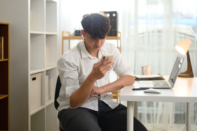 Young Man in White Using Smartphone while Working from Home with Laptop ...