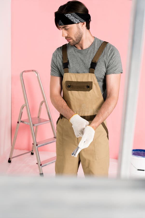 Young Man in Working Overall Putting on Protective Gloves in Room with ...