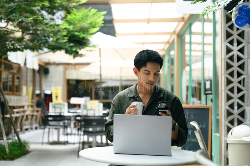 Young Man Working Remotely at an Outdoor Cafe Using Laptop and Checking ...