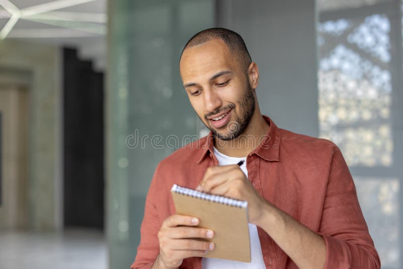 A Young Man is Writing in a Notepad with a Pen, Smiling and Looking ...