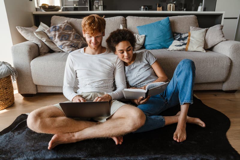 Young Multinational Couple Using Laptop and Reading Book at Home Stock ...