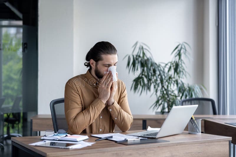 Young Office Worker Dealing with Cold Symptoms at Desk, Using Tissue and Surrounded by Work ...