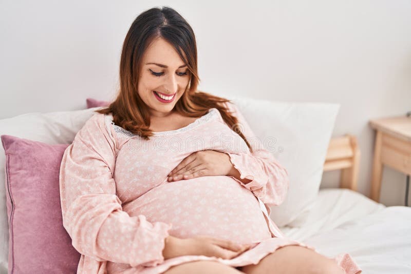 Young Pregnant Woman Touching Belly Sitting On Bed At Bedroom Stock
