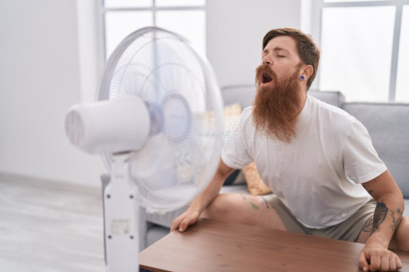 Young Redhead Man Sitting on Sofa Using Fan at Home Stock Photo - Image ...