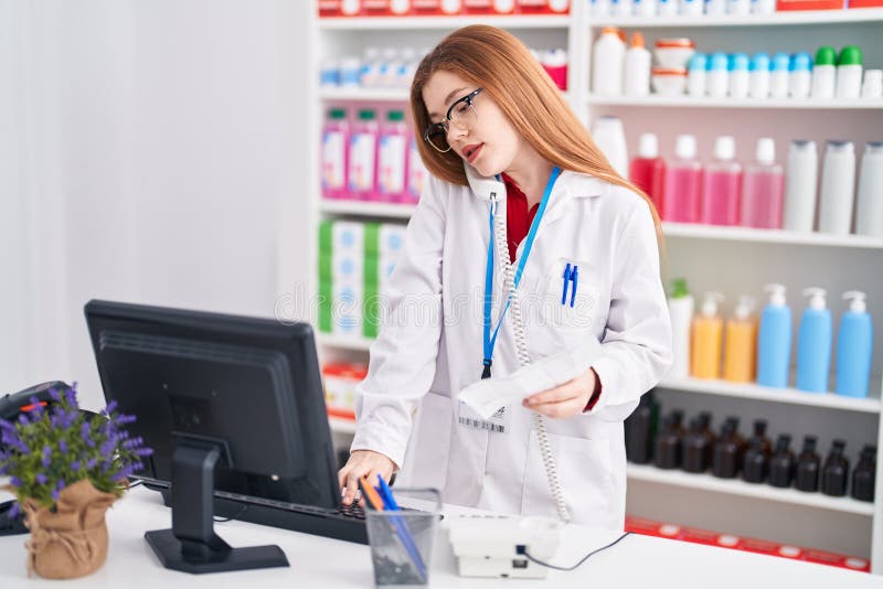 Young Redhead Woman Pharmacist Talking on Telephone Using Computer at ...