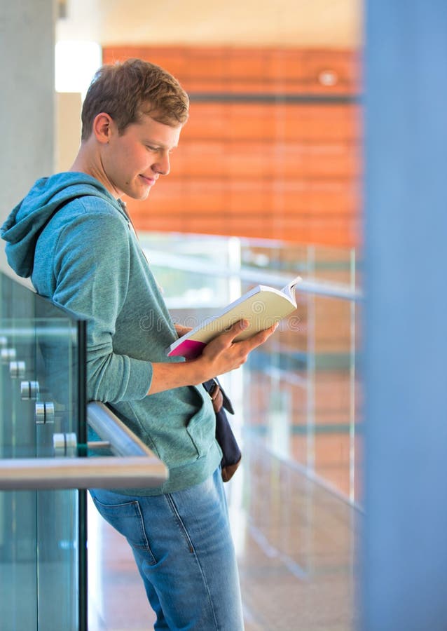 Young Student Reading Book while Standing by Railing at Corridor in ...