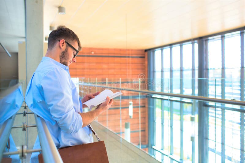 Young Student Reading Book while Standing by Railing at Corridor in ...