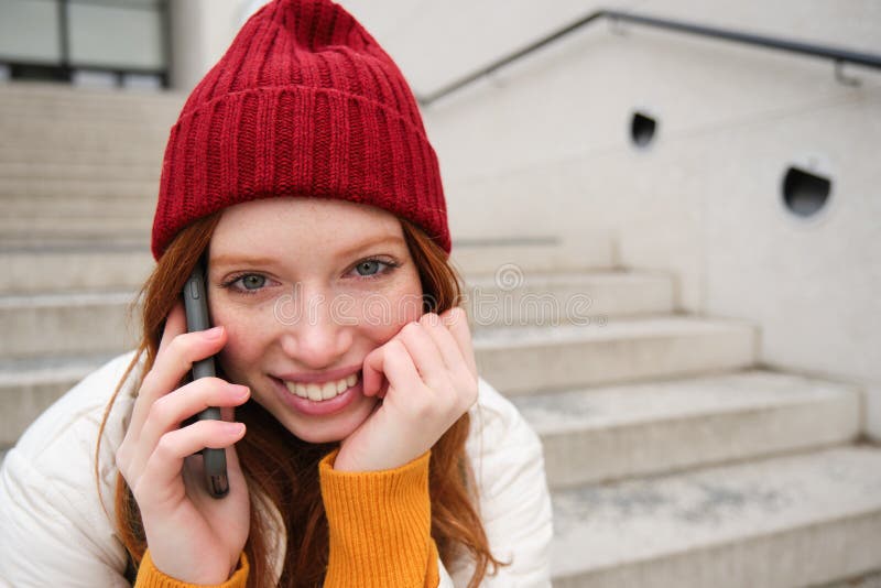 Young Stylish Redhead Girl In Red Hat Sits On Street And Talks On