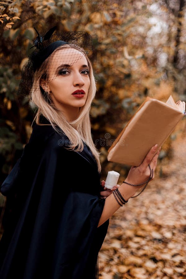 A Young Witch in a Black Cloak Holds a Book with Spells in Her Hands ...