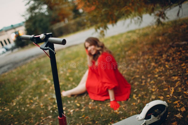 Young Woman Sitting With Electric Scooter In Red Dress At The Autumn