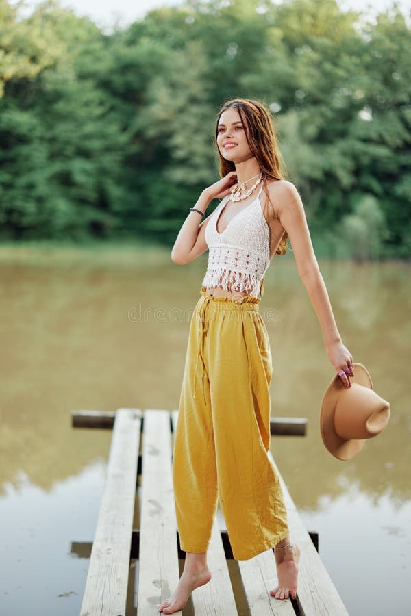 A Young Woman In A Hippie Look And Eco Dress Dancing Outdoors By The Lake Wearing A Hat And
