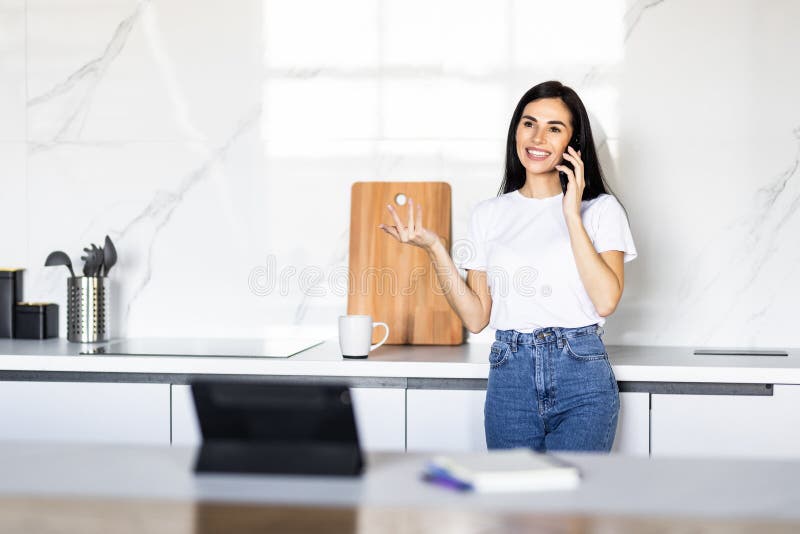Young Woman Sits at the Kitchen Table Using a Laptop and Talking on a ...