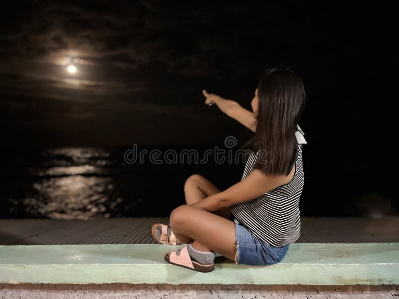 Young Woman Sitting on the Seashore at Night and Pointing at the Moon ...