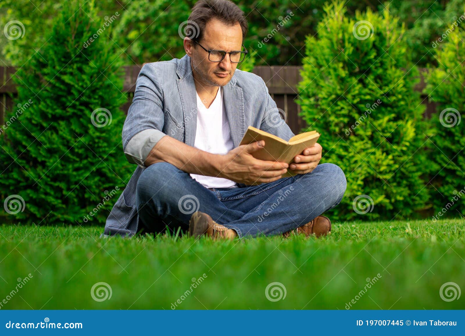 Adult Handsome Man Sitting On Grass In Park With Paper Book Resting