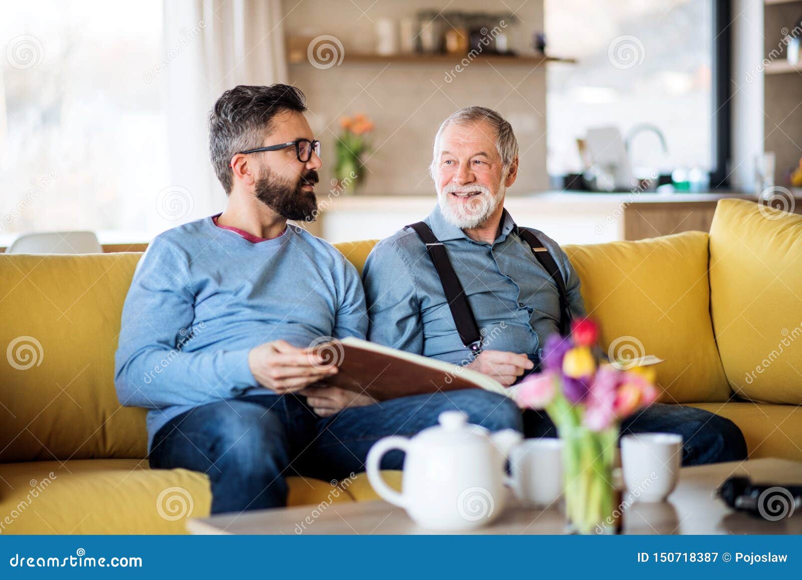 An Adult Hipster Son And Senior Father Sitting On Sofa Indoors At Home