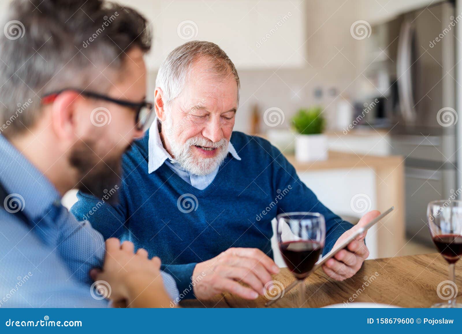 Adult Son And Senior Father Sitting At The Table Indoors At Home Using
