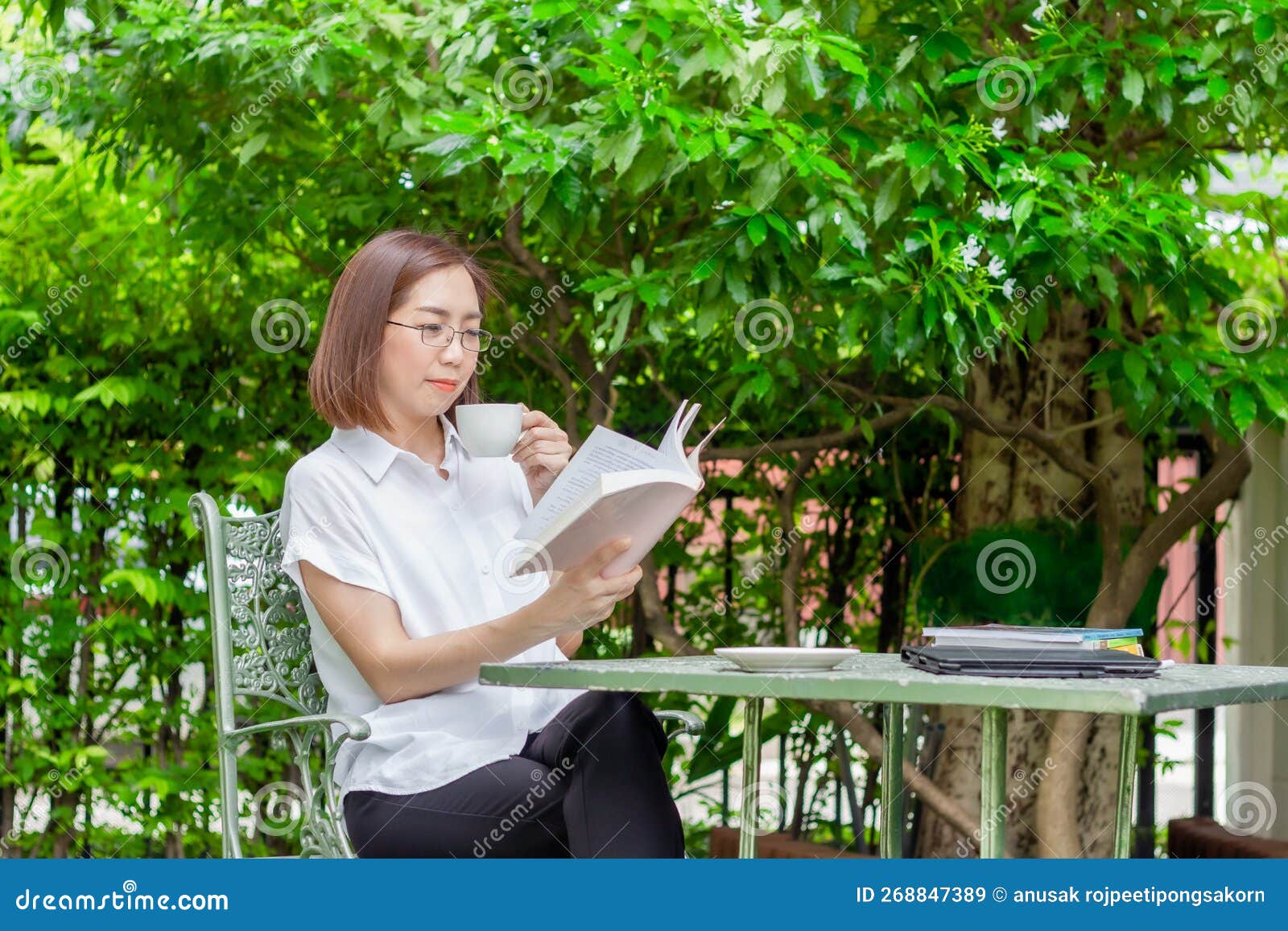 Asian Middle Aged Woman In Glasses Reading A Book In Backyard Stock
