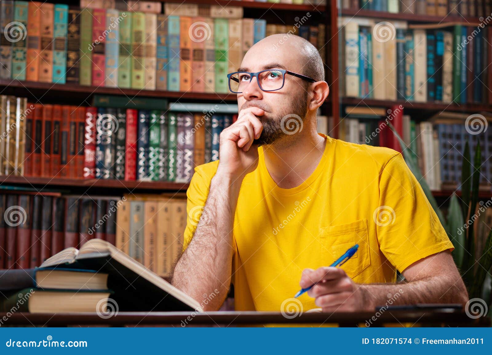 A Bald Bearded Man With Glasses Is Sitting In Front Of His Books