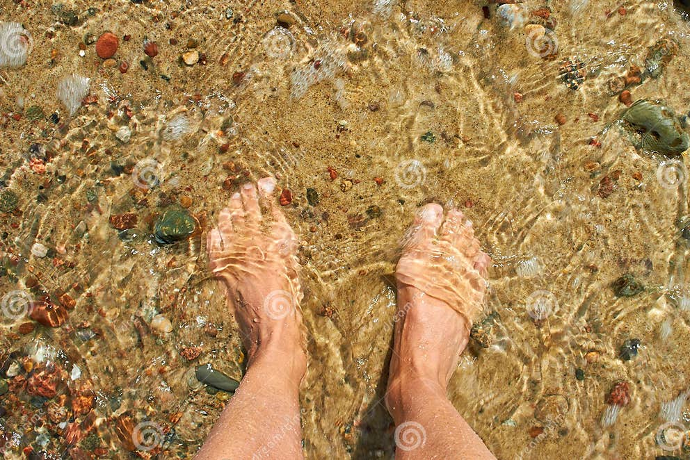 Barefoot Female Feet On A Sandy Beach With Pebbles Washed By A Sea Wave
