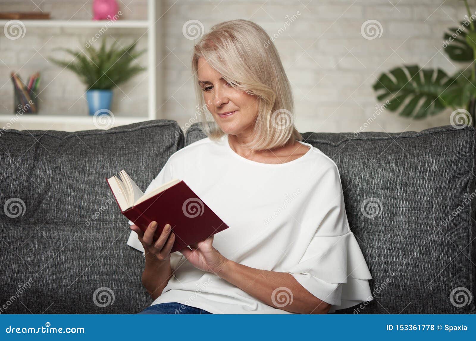 Beautiful Middle Aged Blonde Woman Is Reading A Book At Home Stock