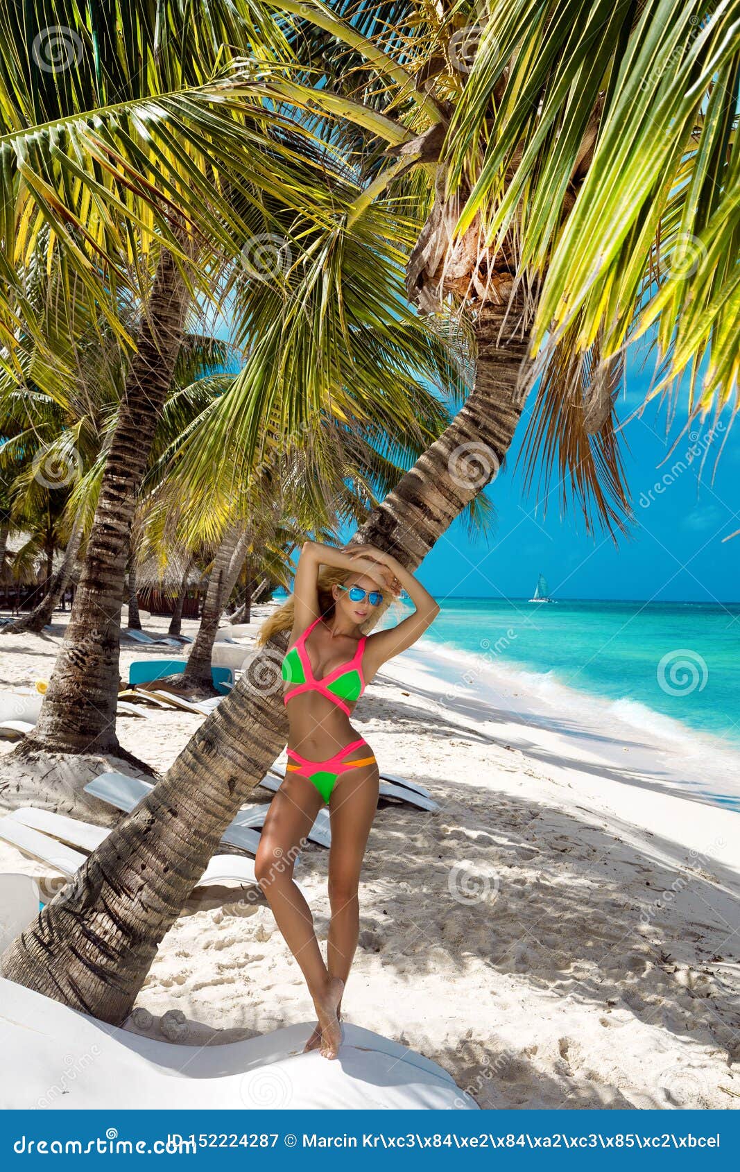 Beautiful Tanned Girl In Fashionable Bikini Stands Next To A Palm Tree