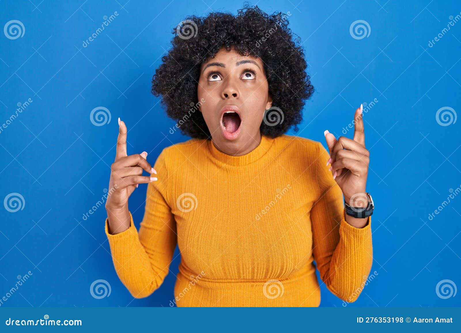 Black Woman With Curly Hair Standing Over Blue Background Amazed And