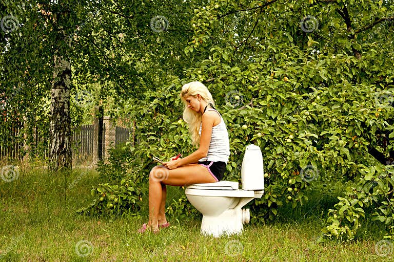 Blond Woman Sitting On A Toilet Bowl And Reading A Book Stock Image