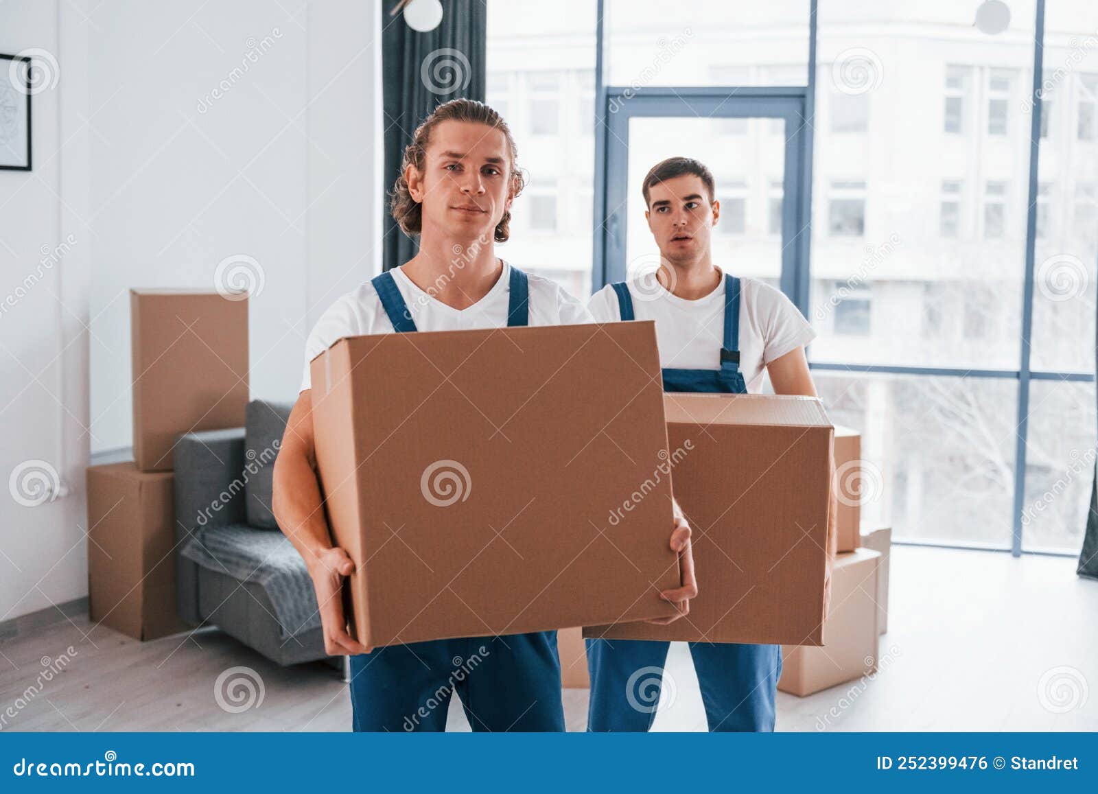 With Boxes in Hands. Two Young Movers in Blue Uniform Working Indoors ...