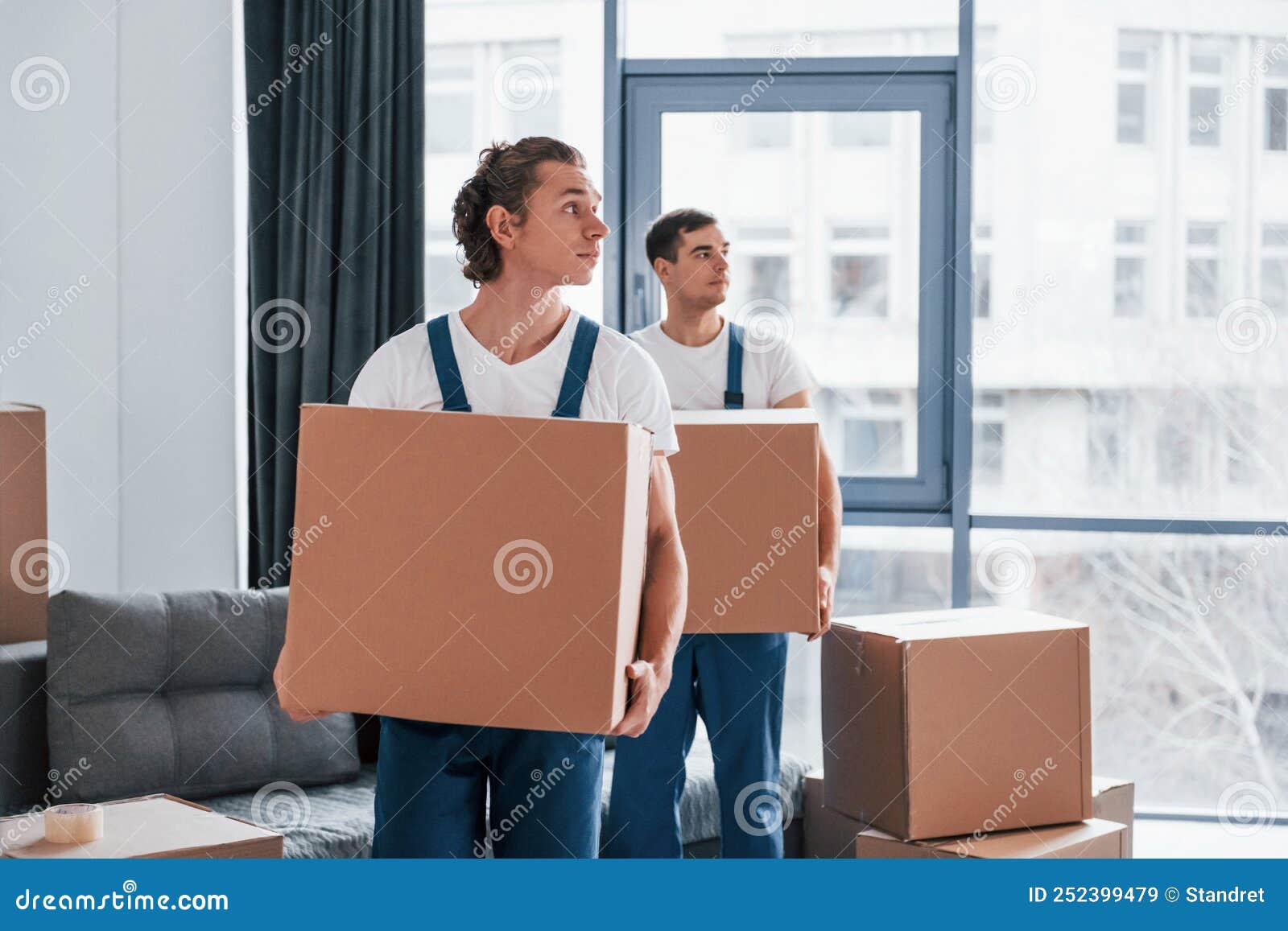 With Boxes in Hands. Two Young Movers in Blue Uniform Working Indoors ...