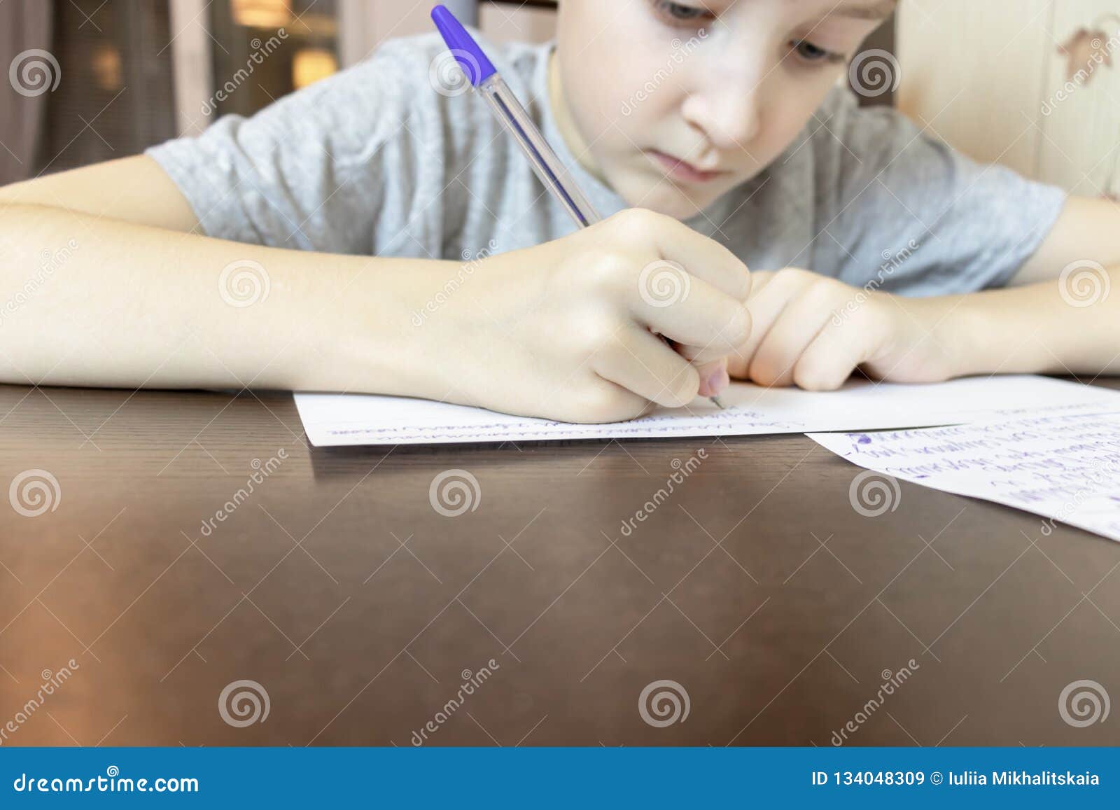 A Boy Sitting by the Table at Home and Writing with a Pen on Paper ...