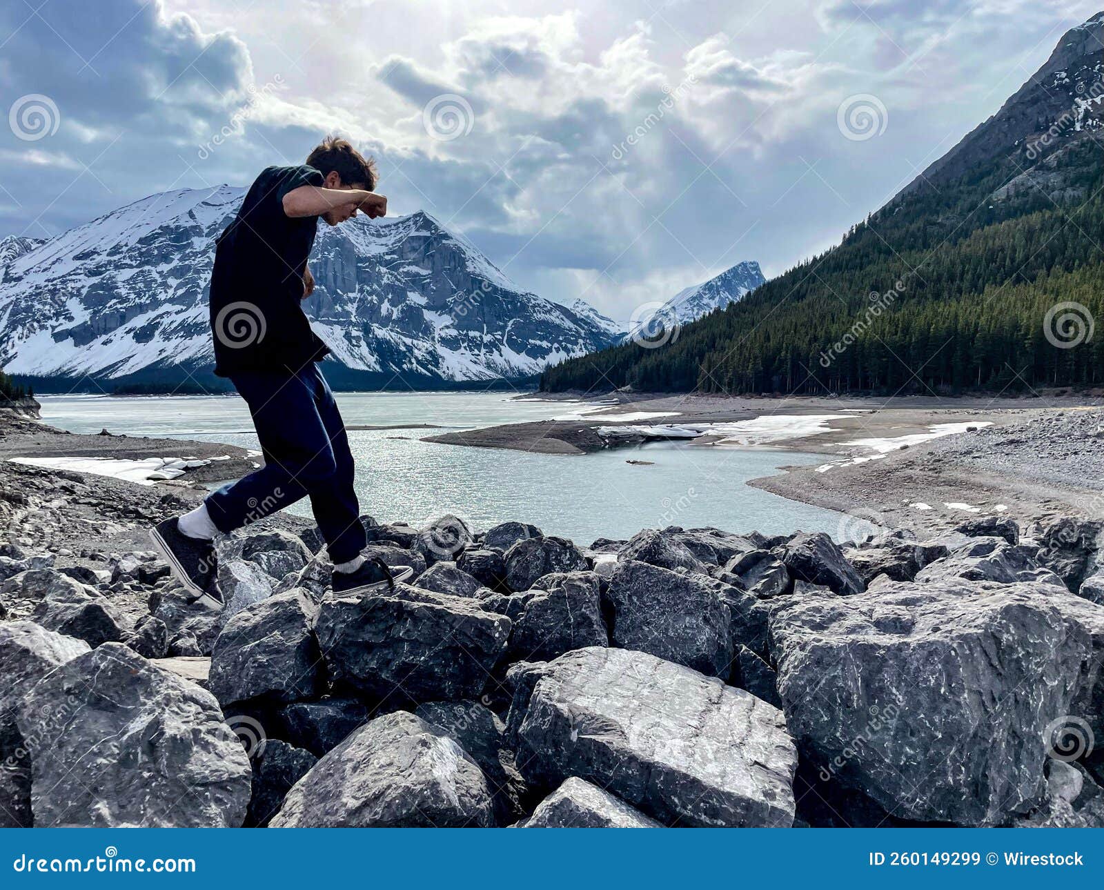 Boy Walking on the Rocks by a Lake with Snowy Mountains in the ...