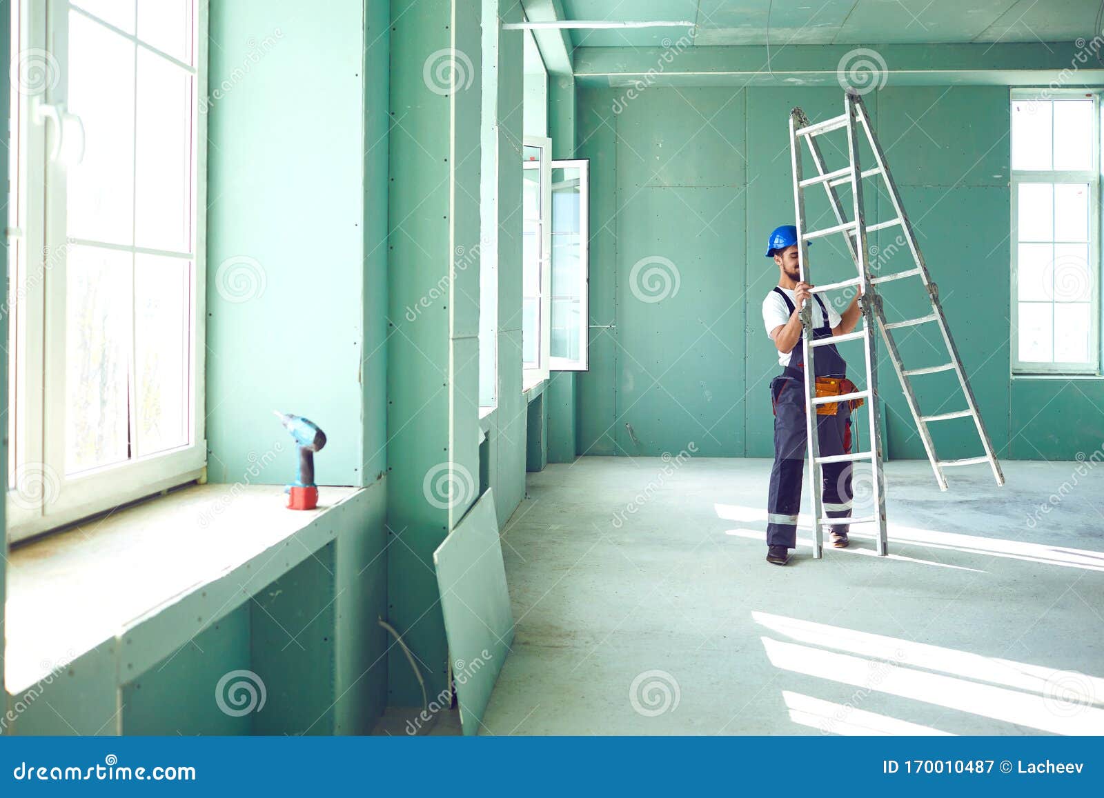A Builder Standing on a Ladder Installs Drywall at a Construction Site ...