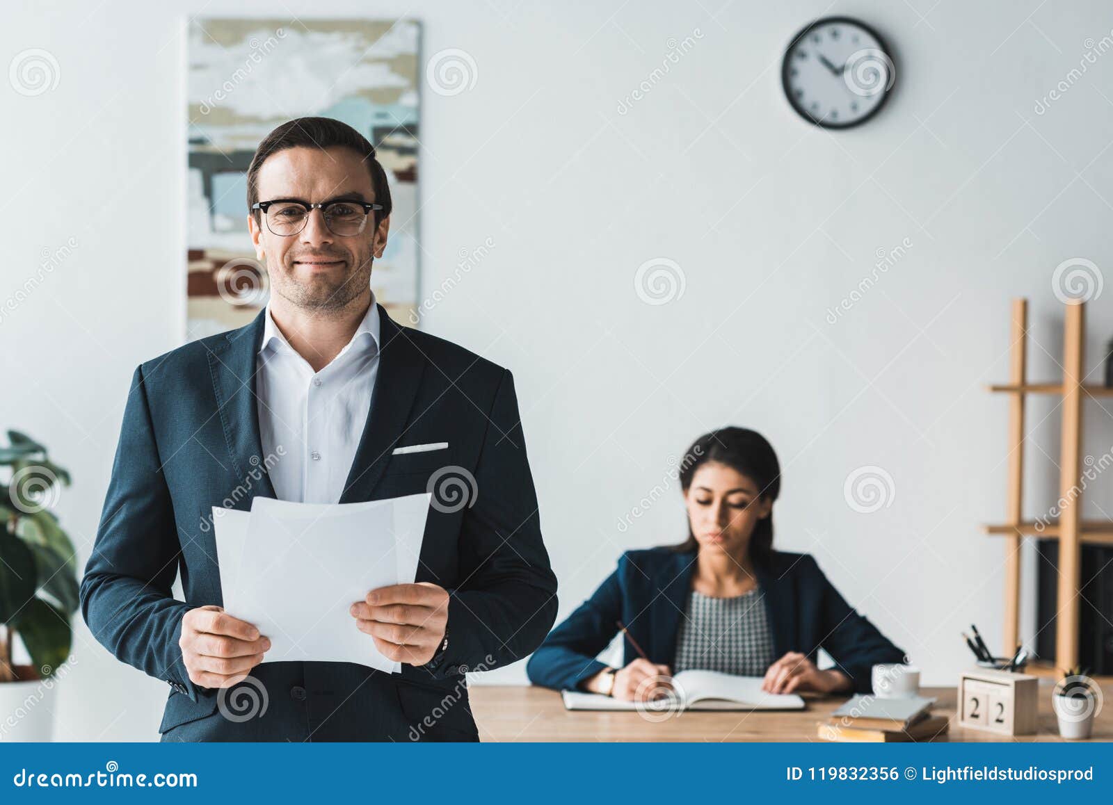 Businessman Holding Papers while Woman Working by Table with Notepad ... Businessman Holding Papers while Woman Working by Table with Notepad ...