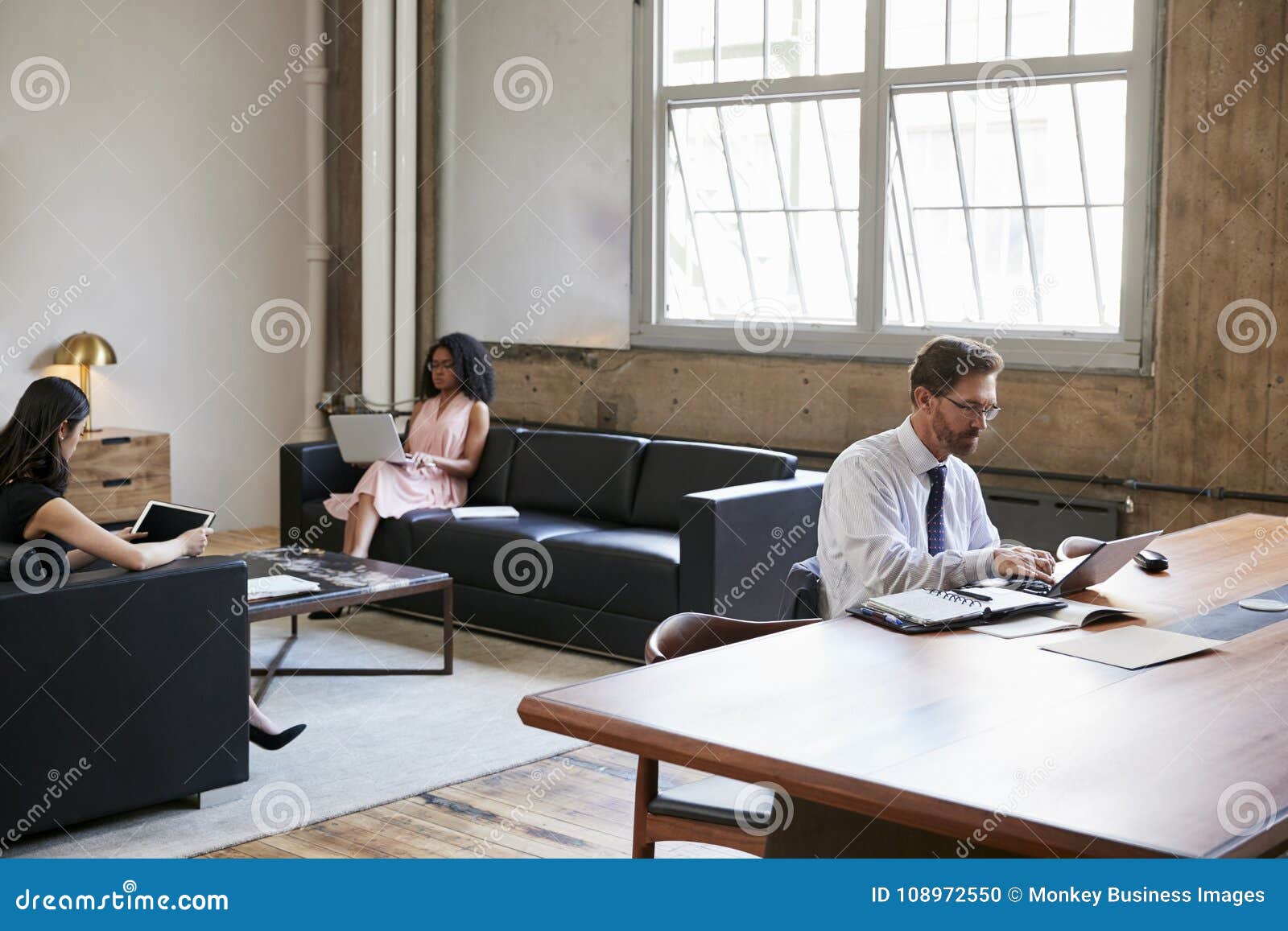 Businessman Working at Desk, Colleagues on Sofas, Side View Stock Photo ...