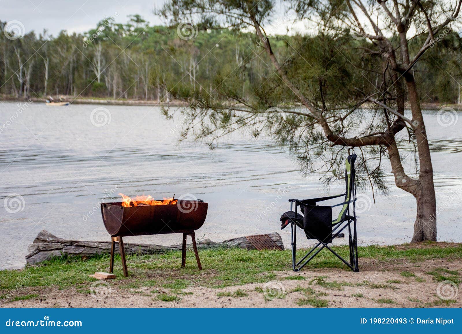 Campfire in a Half Barrel Fire Pit and Empty Campchair on the Lake ...
