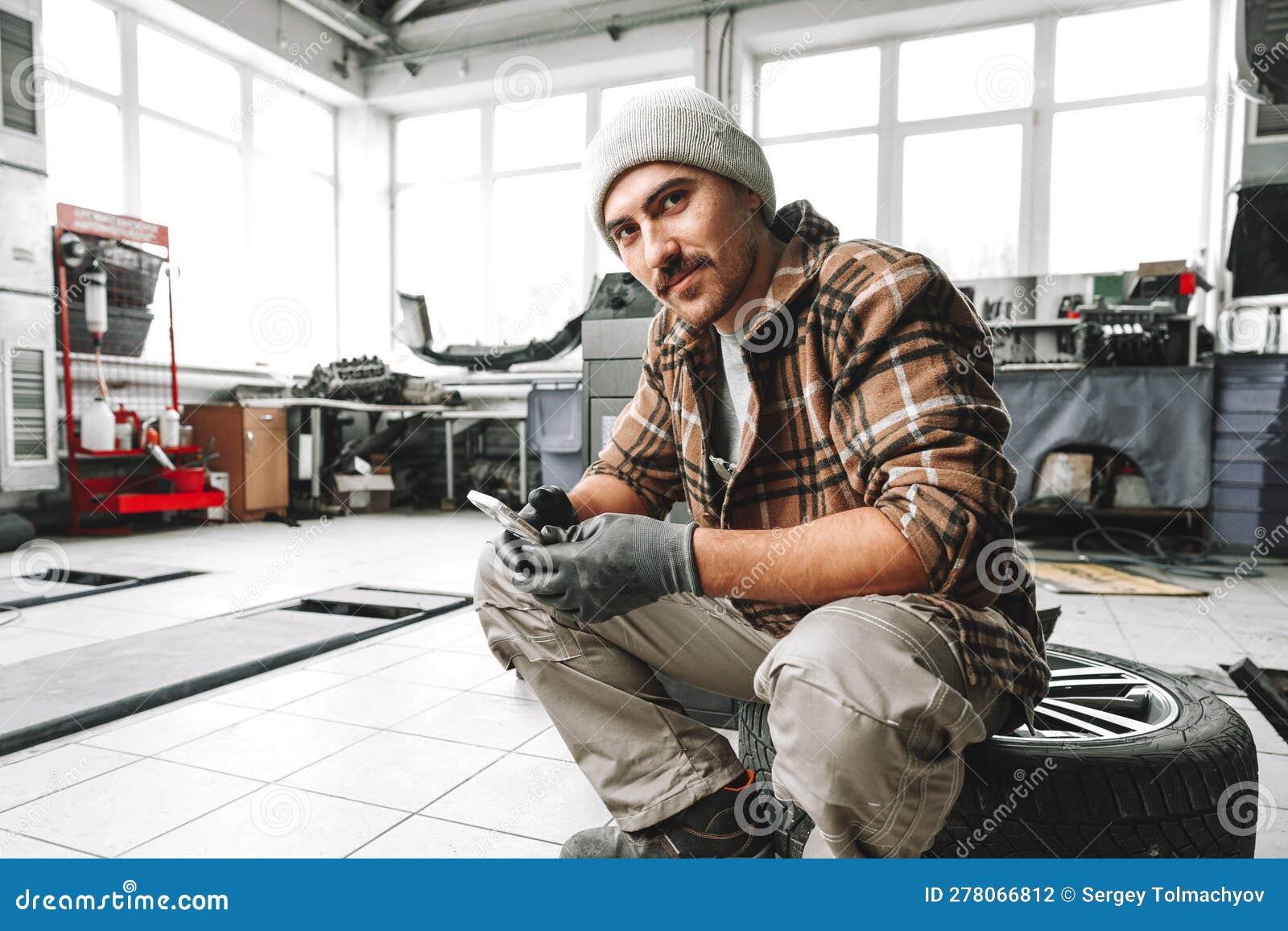 Car Service Worker Sitting on a Wheel and Resting, Using Mobile Phone ...