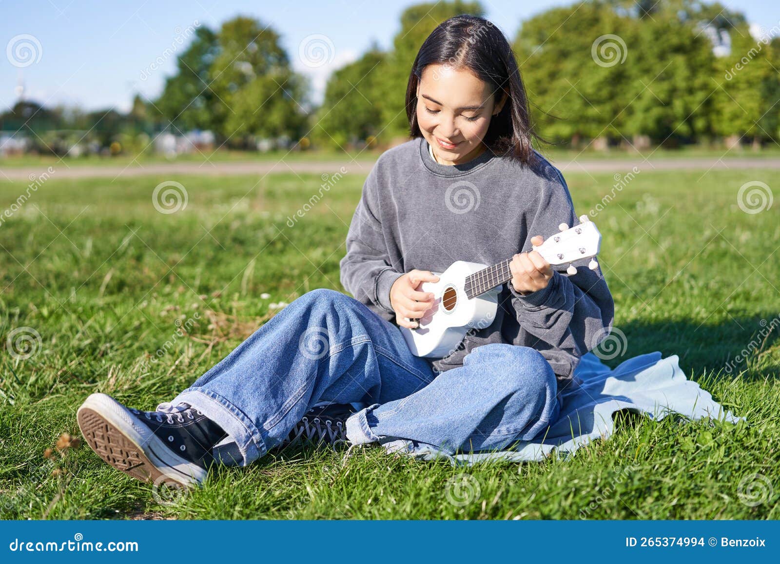 Carefree Asian Girl Singing And Playing Ukulele In Park Sitting On