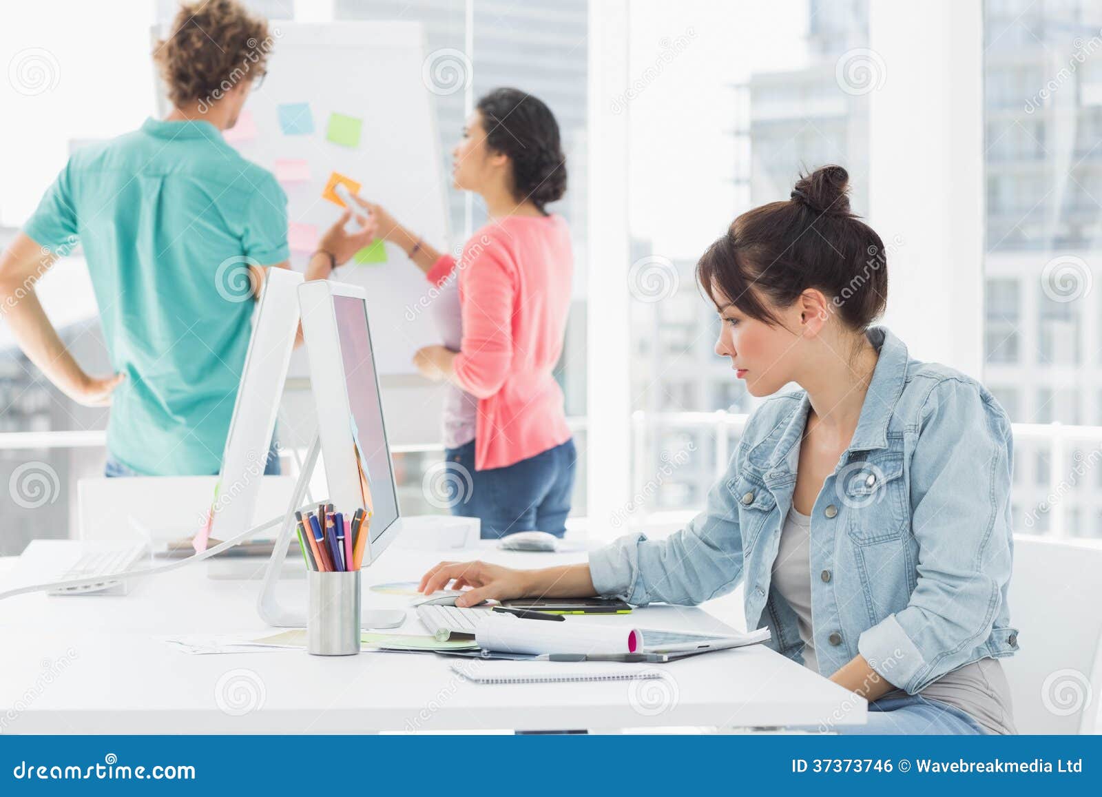 Casual Woman Using Computer with Colleagues Behind in Office Stock ...