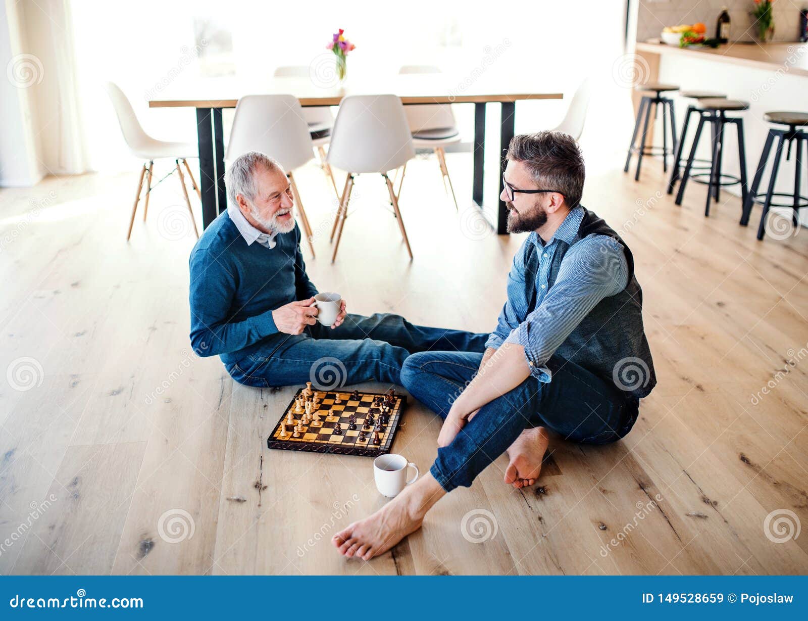 An Adult Hipster Son And Senior Father Sitting On Floor Indoors At Home