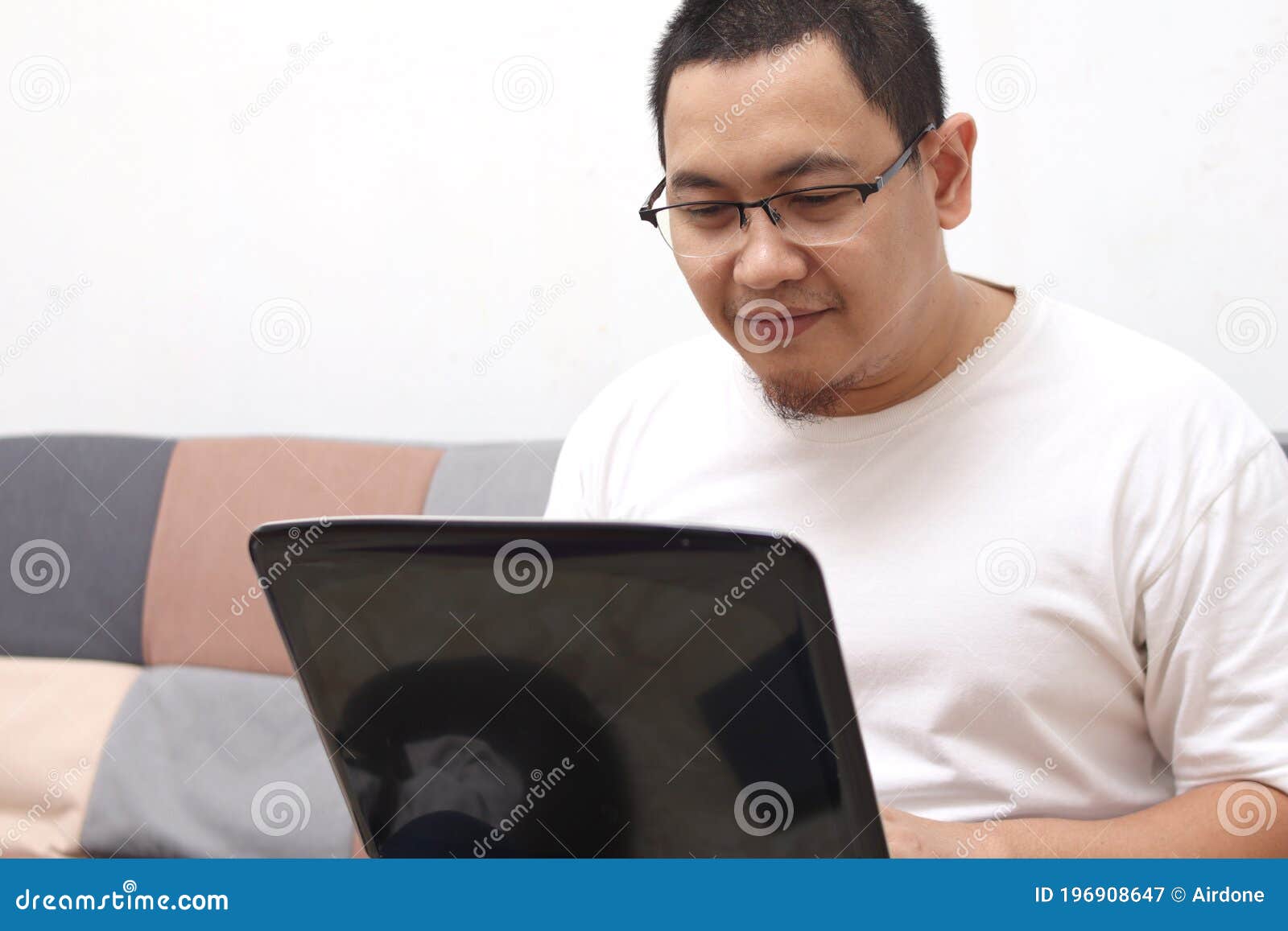 Cheerful Asian Man Using Computer and Smiling while Sitting on the Sofa ...