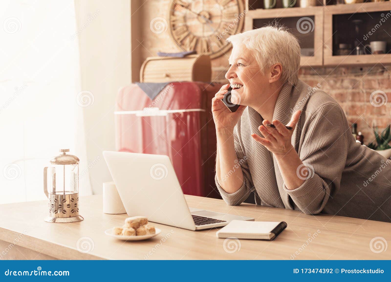 Cheerful Elderly Woman Talking on Cellphone and Using Laptop in Kitchen ...