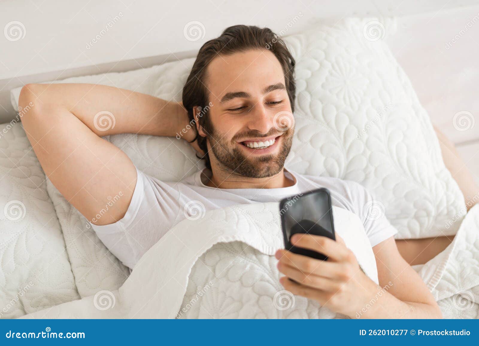 Cheerful Millennial European Man With Stubble Lies On White Bed Typing